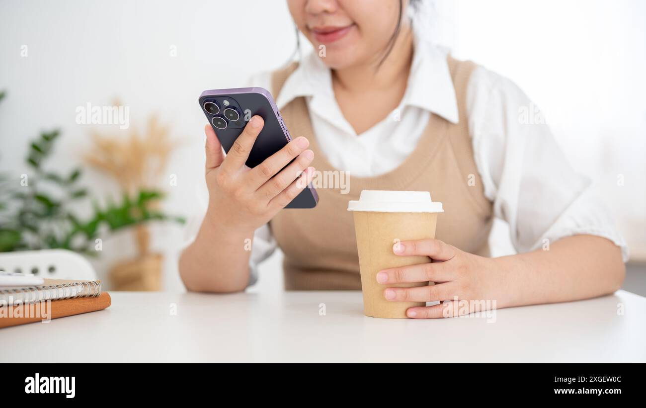 A cropped image of a happy Asian woman relaxing in a coffee shop, using her smartphone and ...