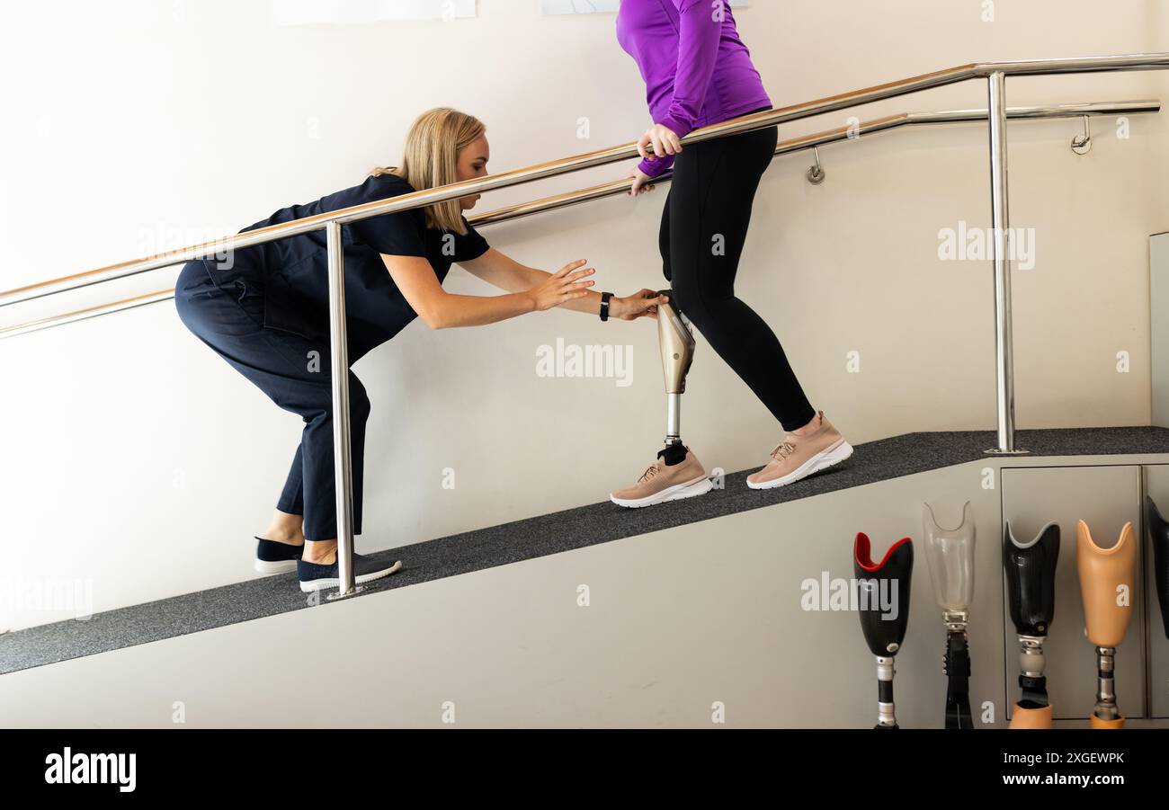Therapist assisting woman with prosthetic leg walking down ramp in clinic. Rehabilitation ...