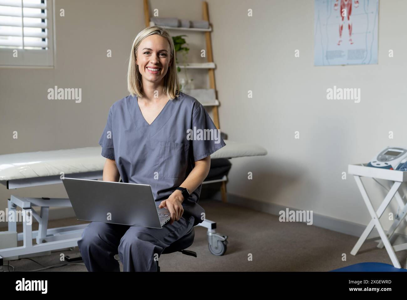 Smiling nurse in scrubs using laptop in medical office, sitting on ...