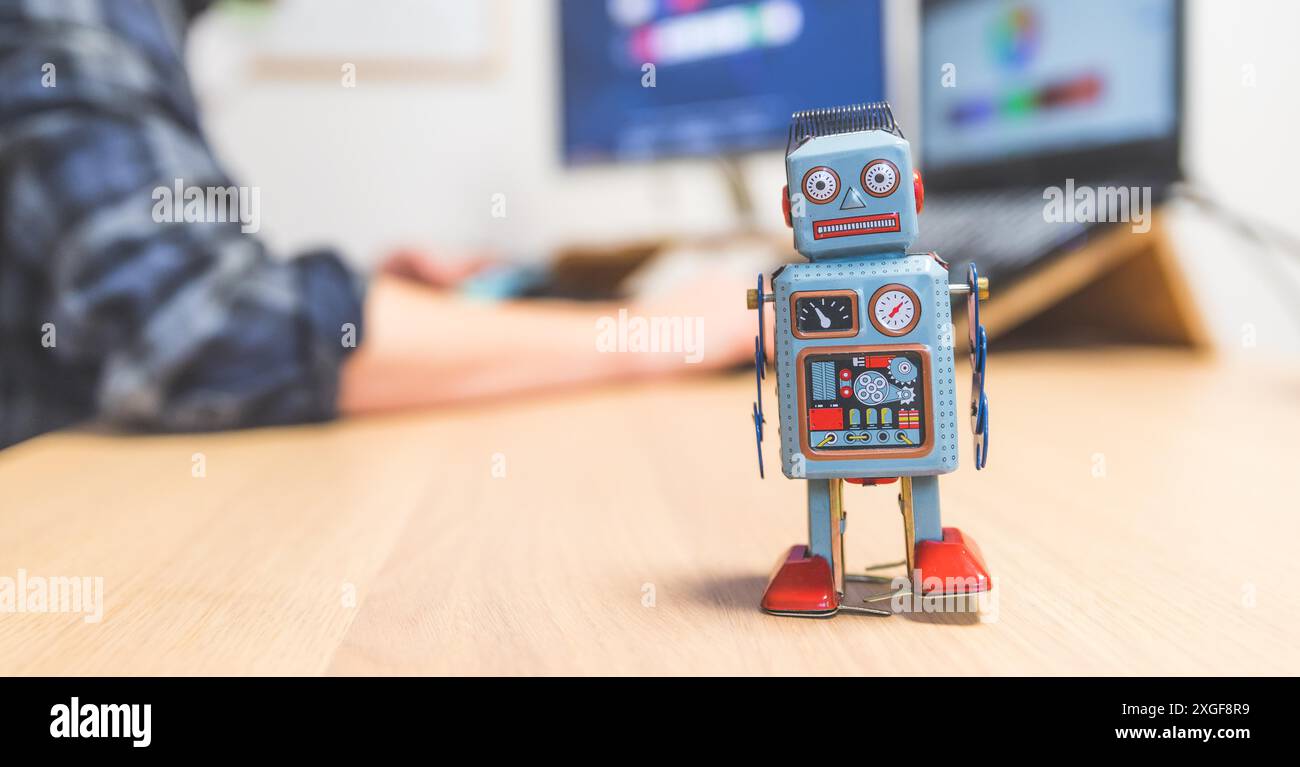 Toy robot on a desktop, man working on the computer in the background. Symbol for artificial ...