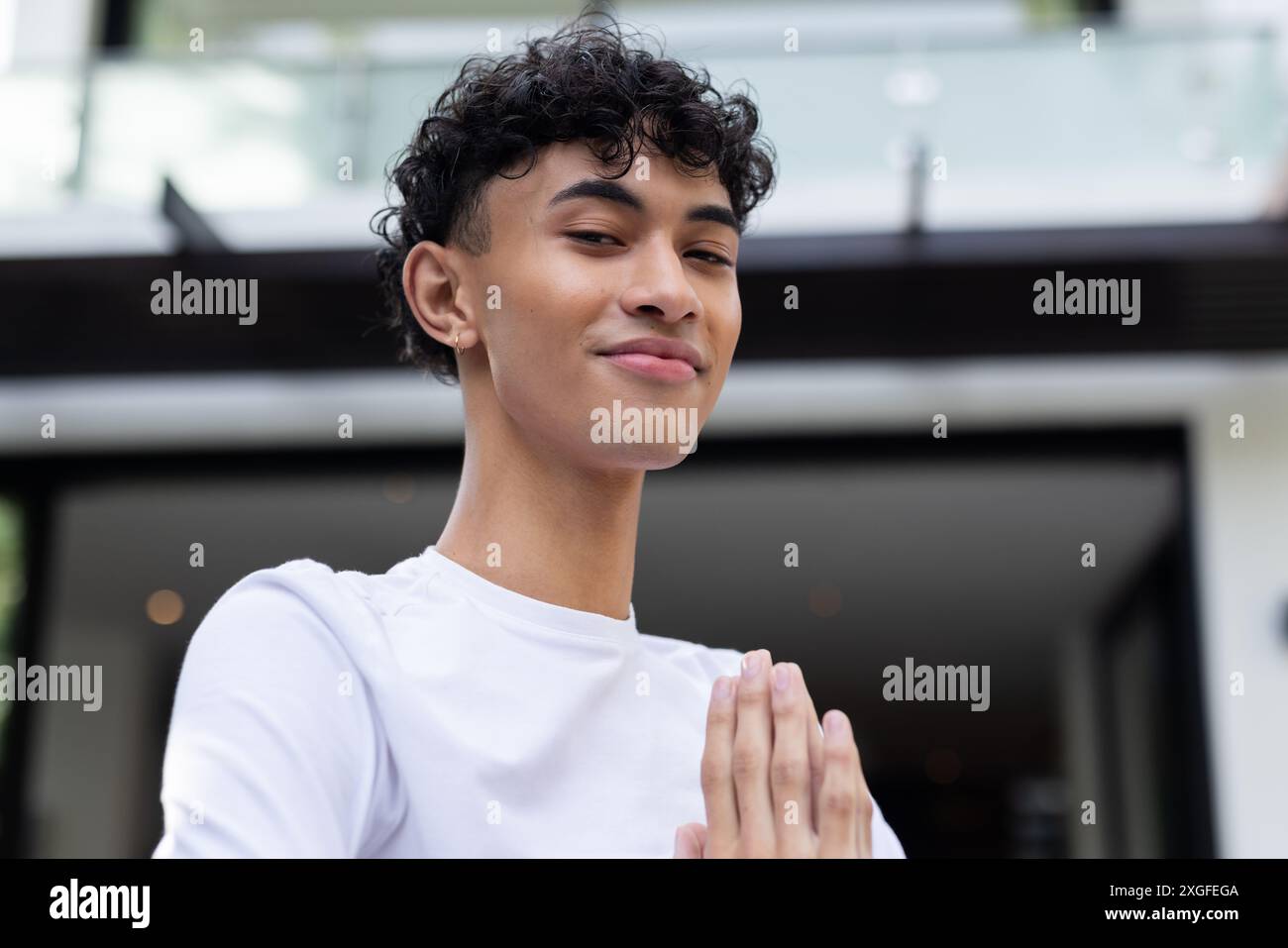 Smiling young man with hands in prayer pose, standing outside modern ...