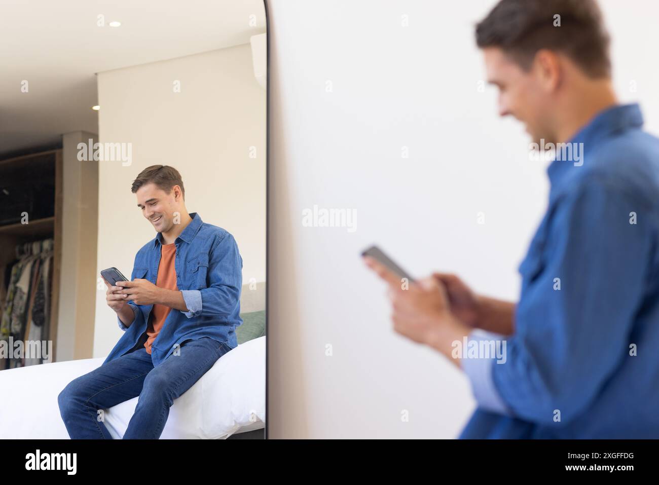 Using smartphone, man sitting on bed and smiling at reflection in ...