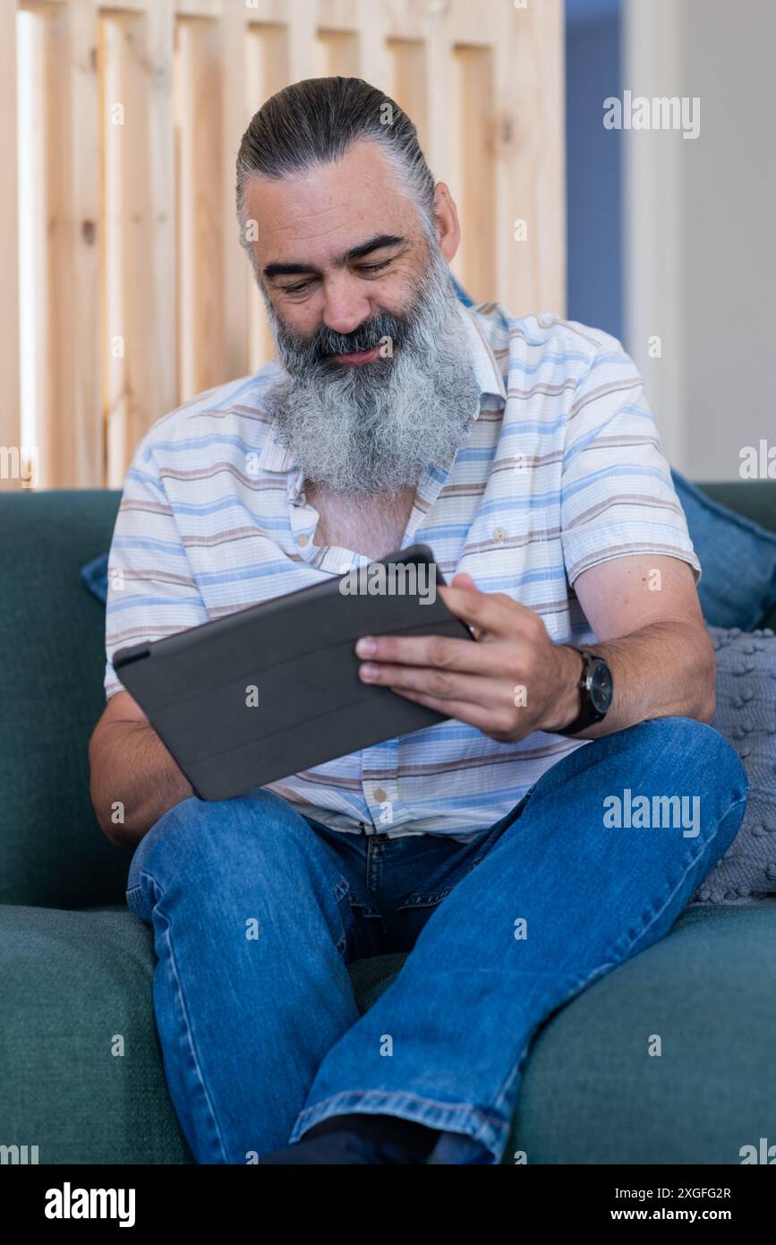 Using tablet, senior man with gray beard sitting on couch, smiling and ...