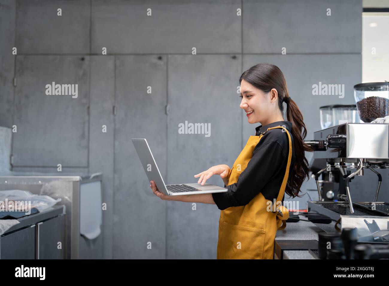 Young Female Barista Using Laptop in Modern Coffee Shop Wearing Yellow Apron and Smiling Stock ...