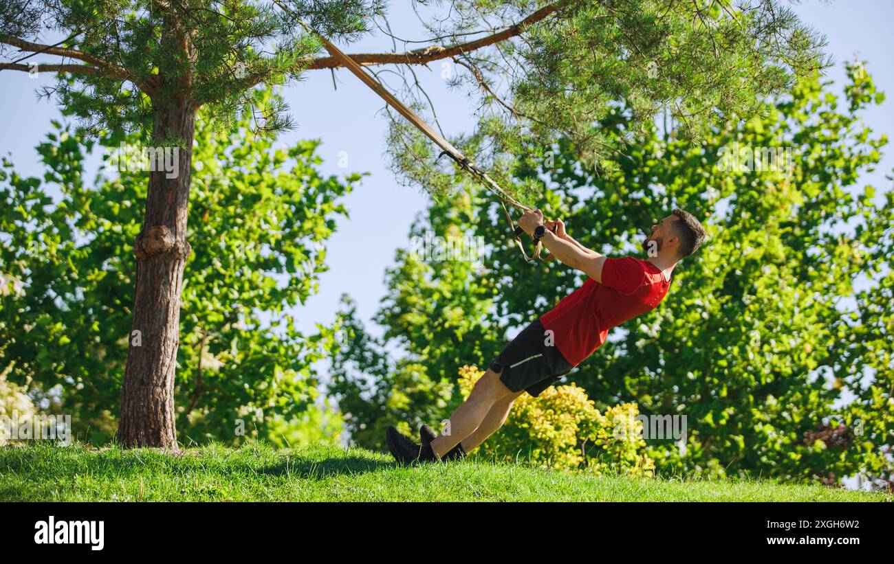 Muscular man doing TRX workout in lush green park, using tree branches ...