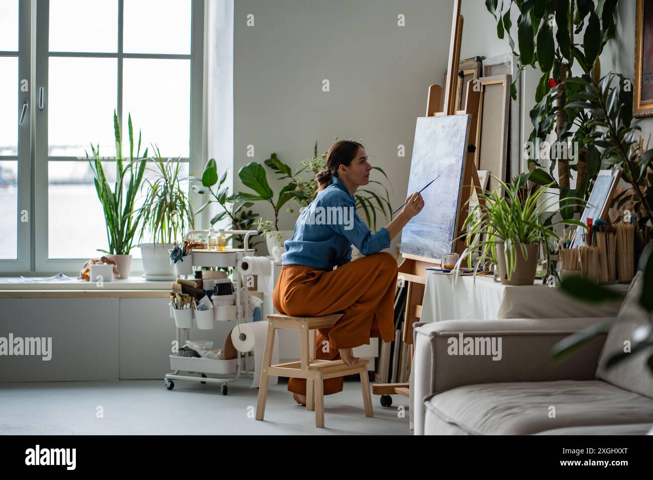 Focused woman artist paints in oil, sits with brush, looks at canvas with painting in art studio ...