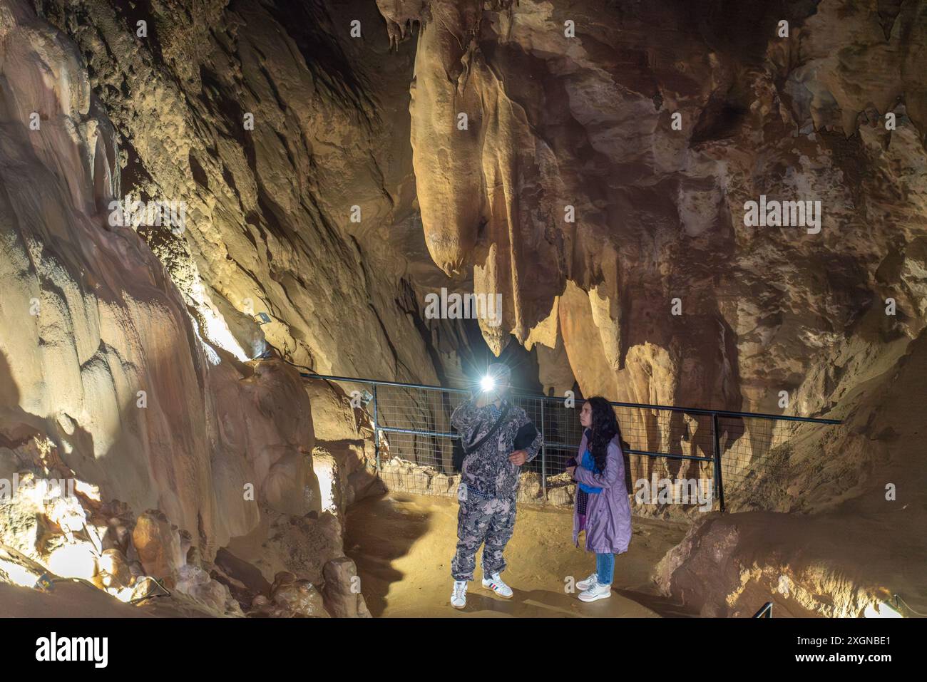 Two people are walking through a cave with a light on. The cave is dark ...