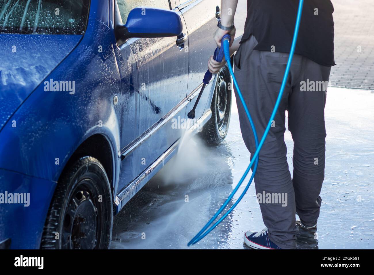A man is washing a blue car with a hose in the driveway. The cars shiny ...