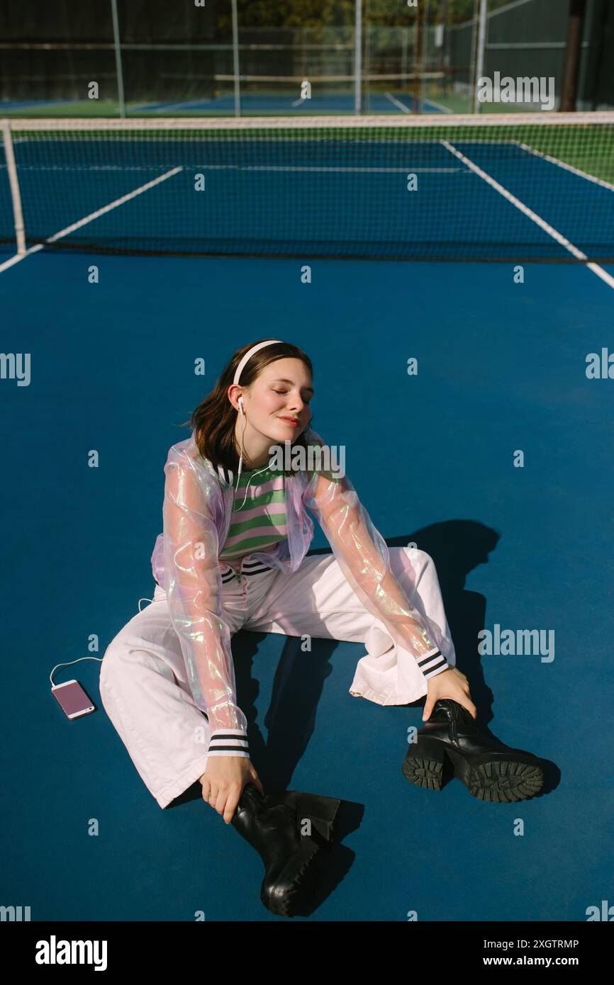 A young woman sits relaxed on a blue tennis court, listening to music ...
