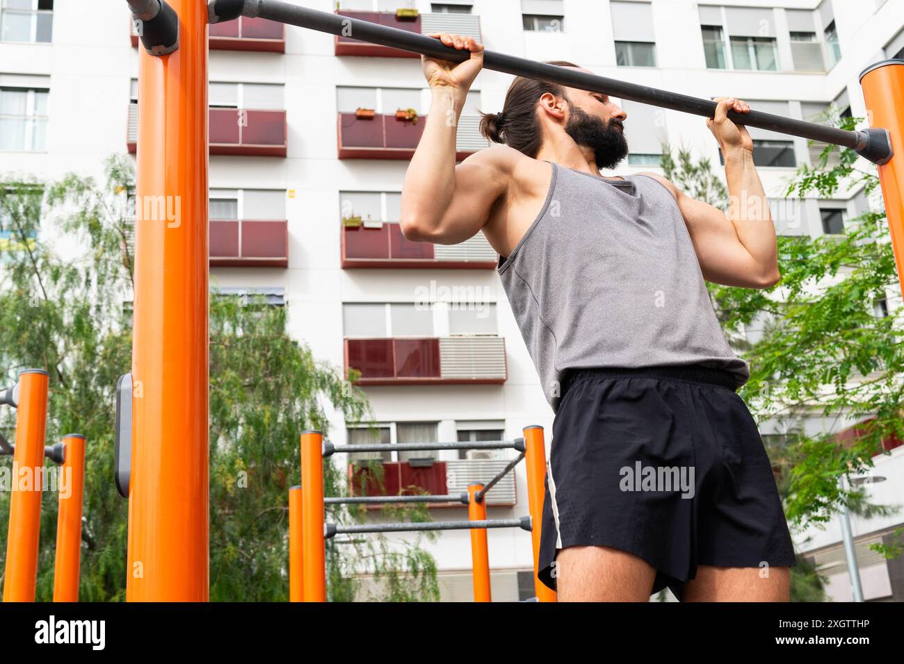 A young, bearded man exercises on an orange pull-up bar in an outdoor ...