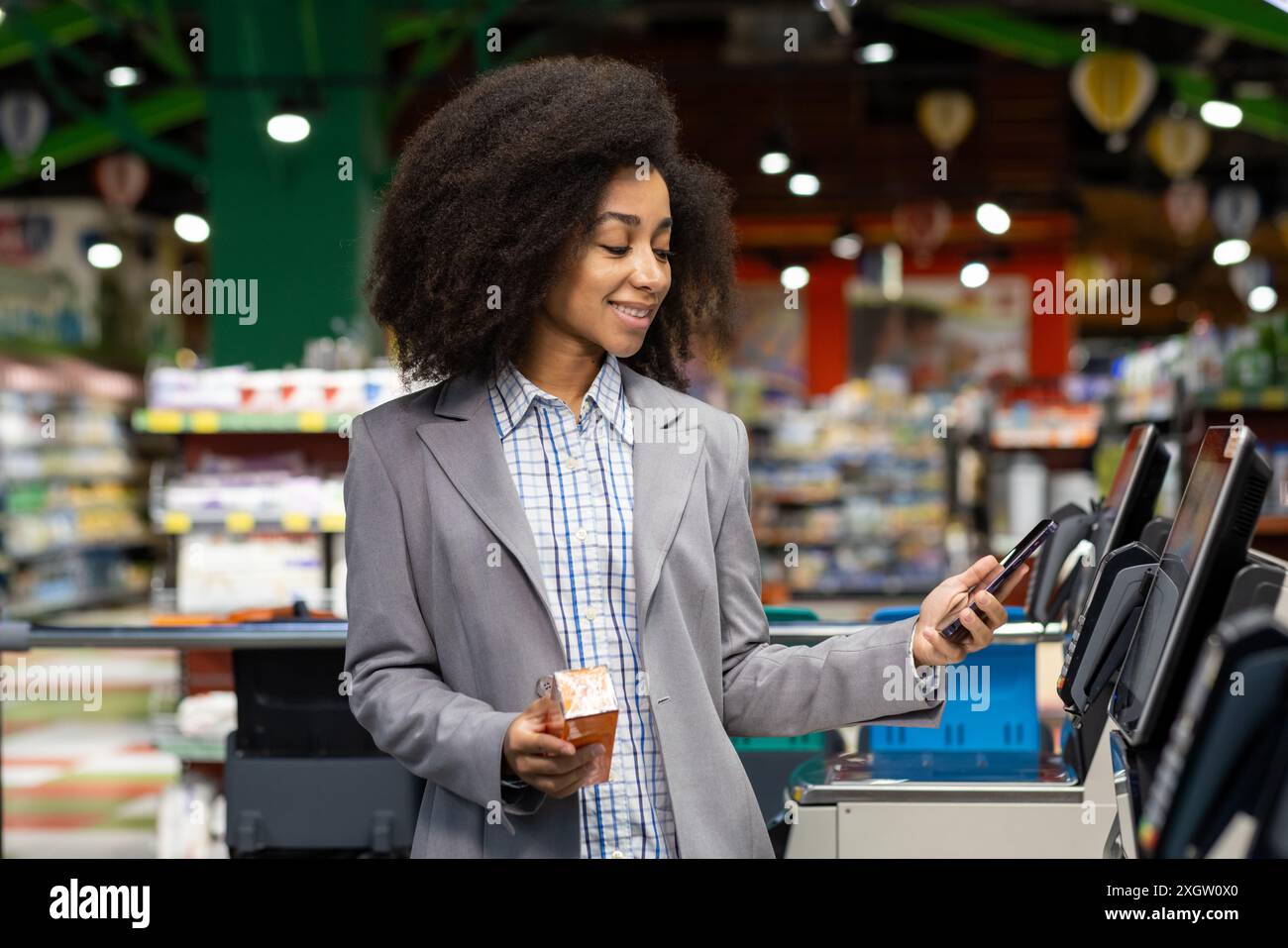 Smiling woman using smartphone at self-checkout kiosk in grocery store. Professional attire ...
