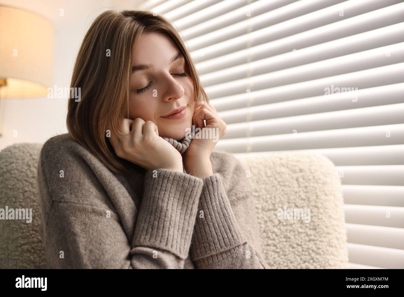 Woman sitting on armchair near window blinds at home, space for text ...