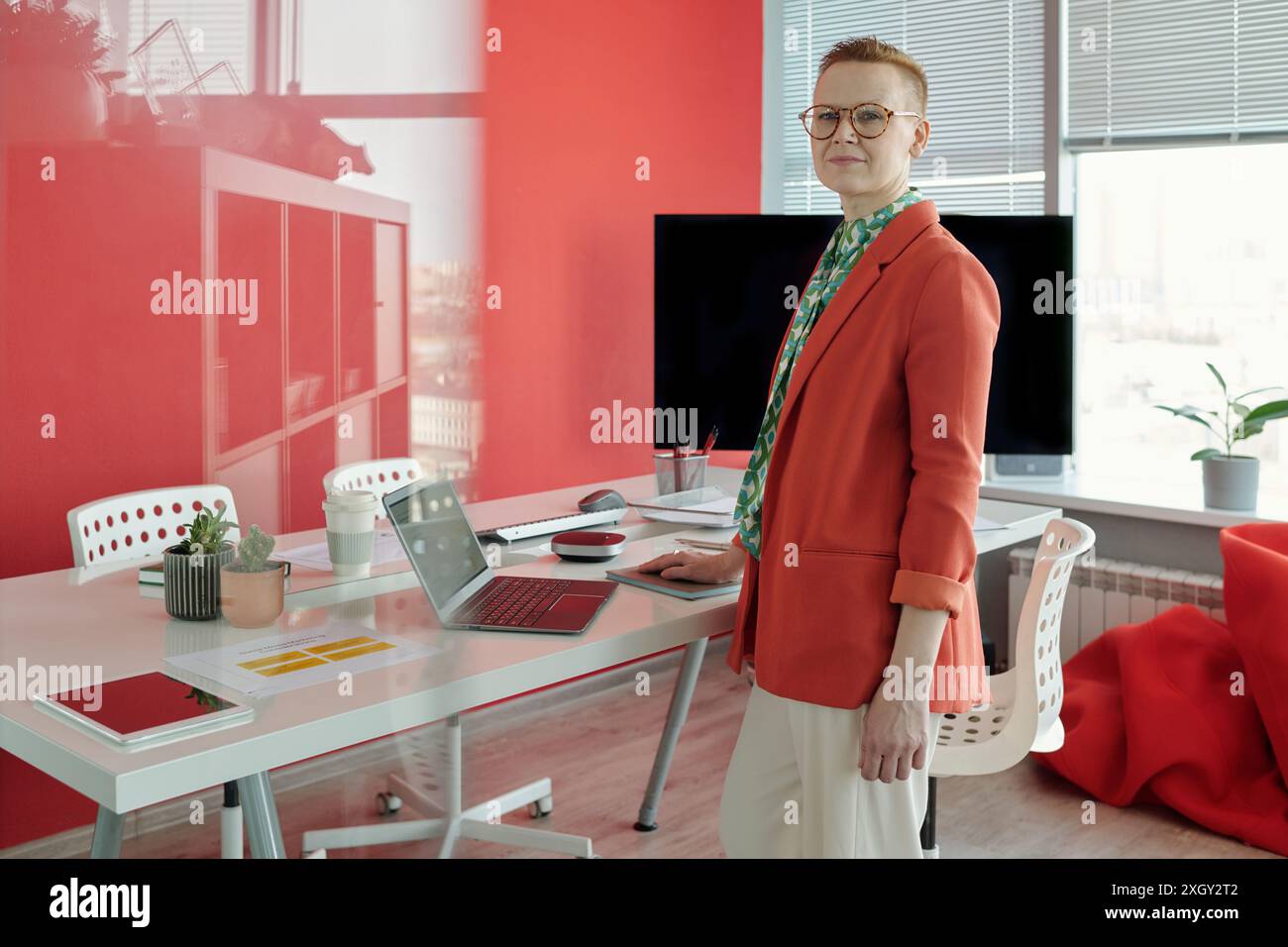 Portrait of female office worker standing by desk with laptop in bright ...