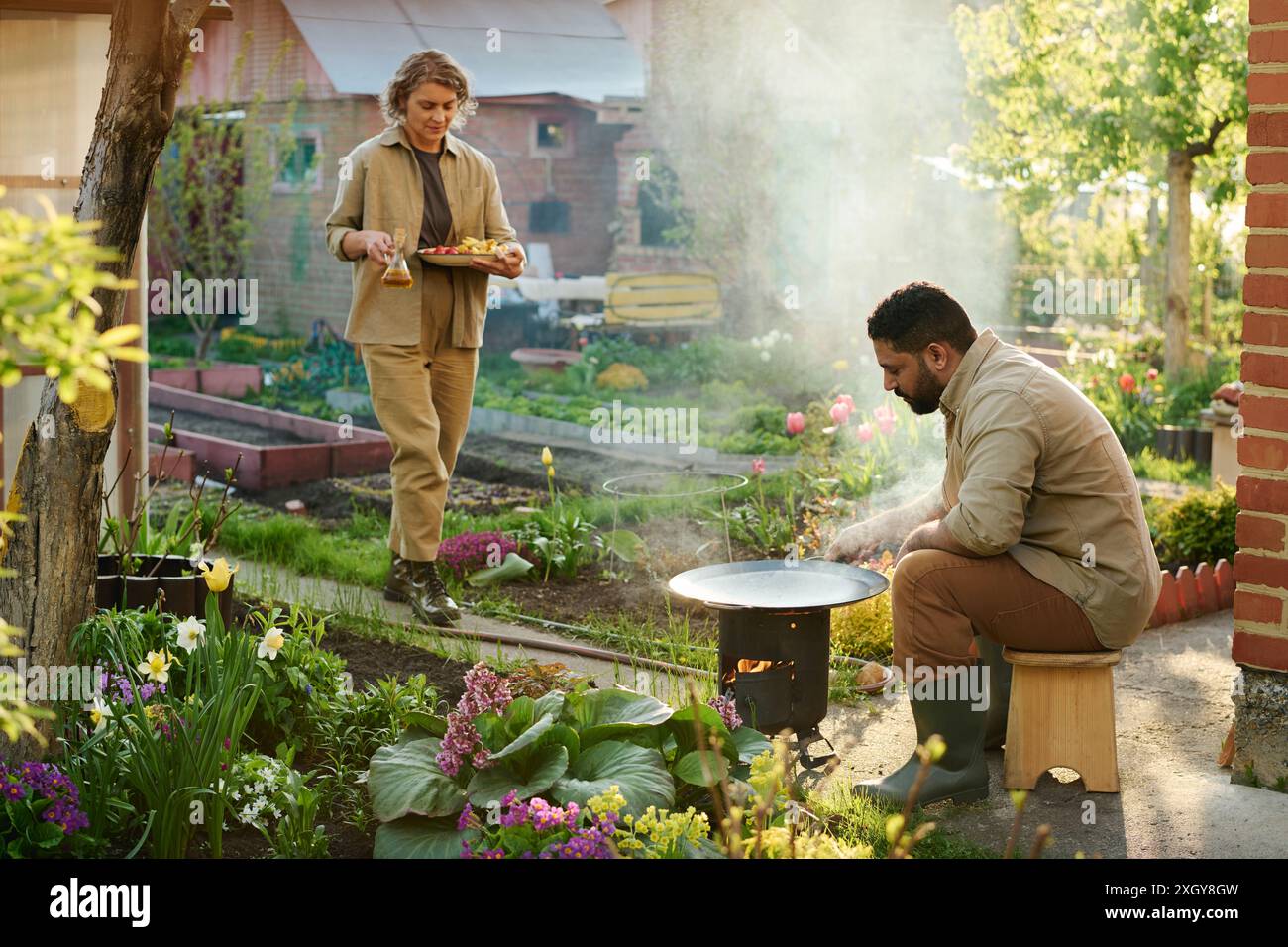 Mature man bending over frying pan on iron barrel smoker while his wife ...