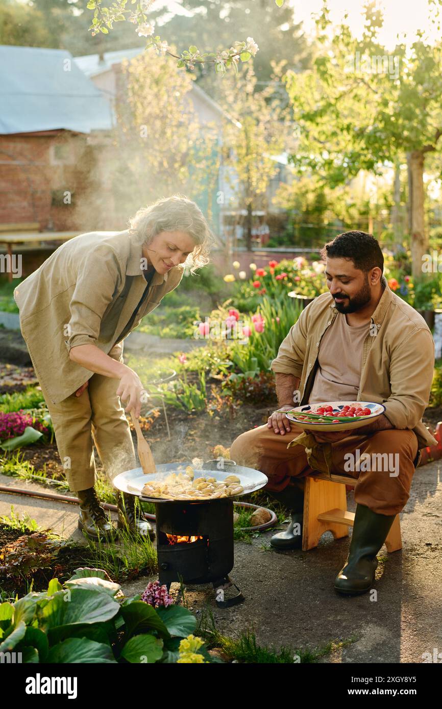 Happy woman bending over frying pan and mixing chopped potatoes while ...