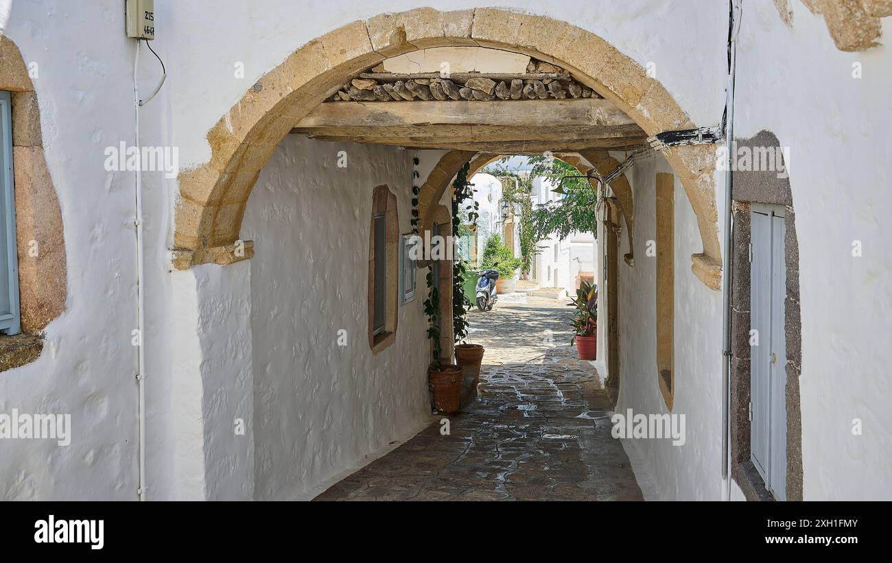 A shady passageway through a stone archway, flanked by white buildings ...