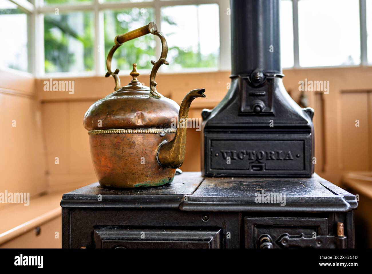 Rustic vintage brass kettle on an antique wood-burning stove in a cosy, sunlit room with large ...