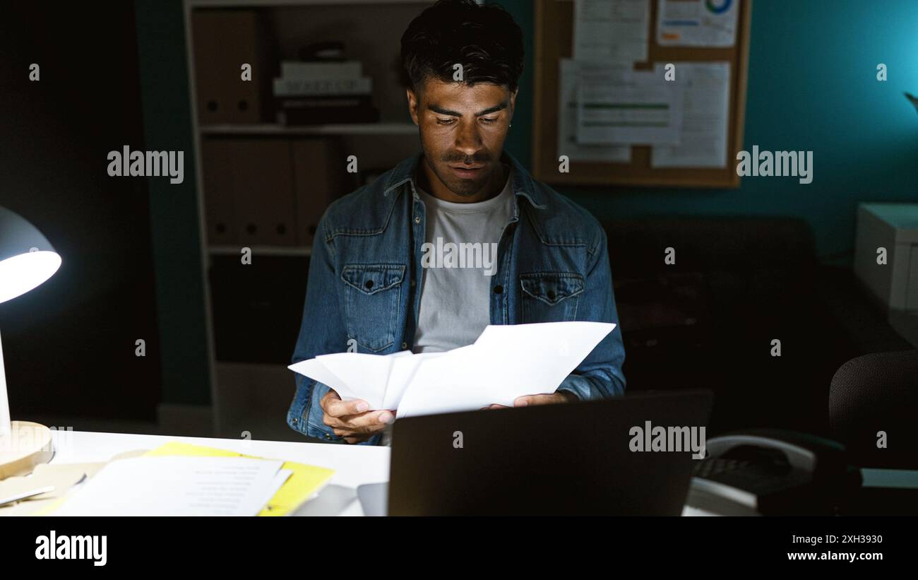 A focused hispanic man with a beard reviews documents at night in a ...