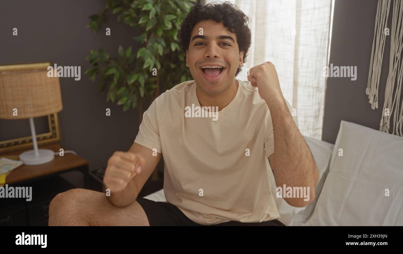 A handsome hispanic man happily sits on a bed in a cozy bedroom with ...