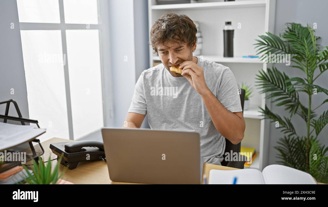 A bearded man snacking while working on a laptop in a modern home ...