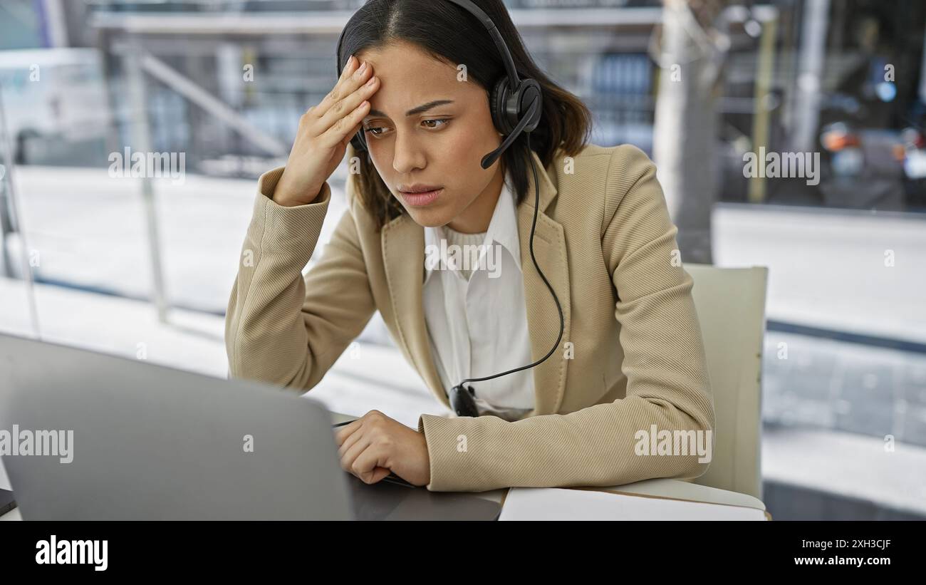 Stressed young hispanic woman in a headset sits at her desk with a ...