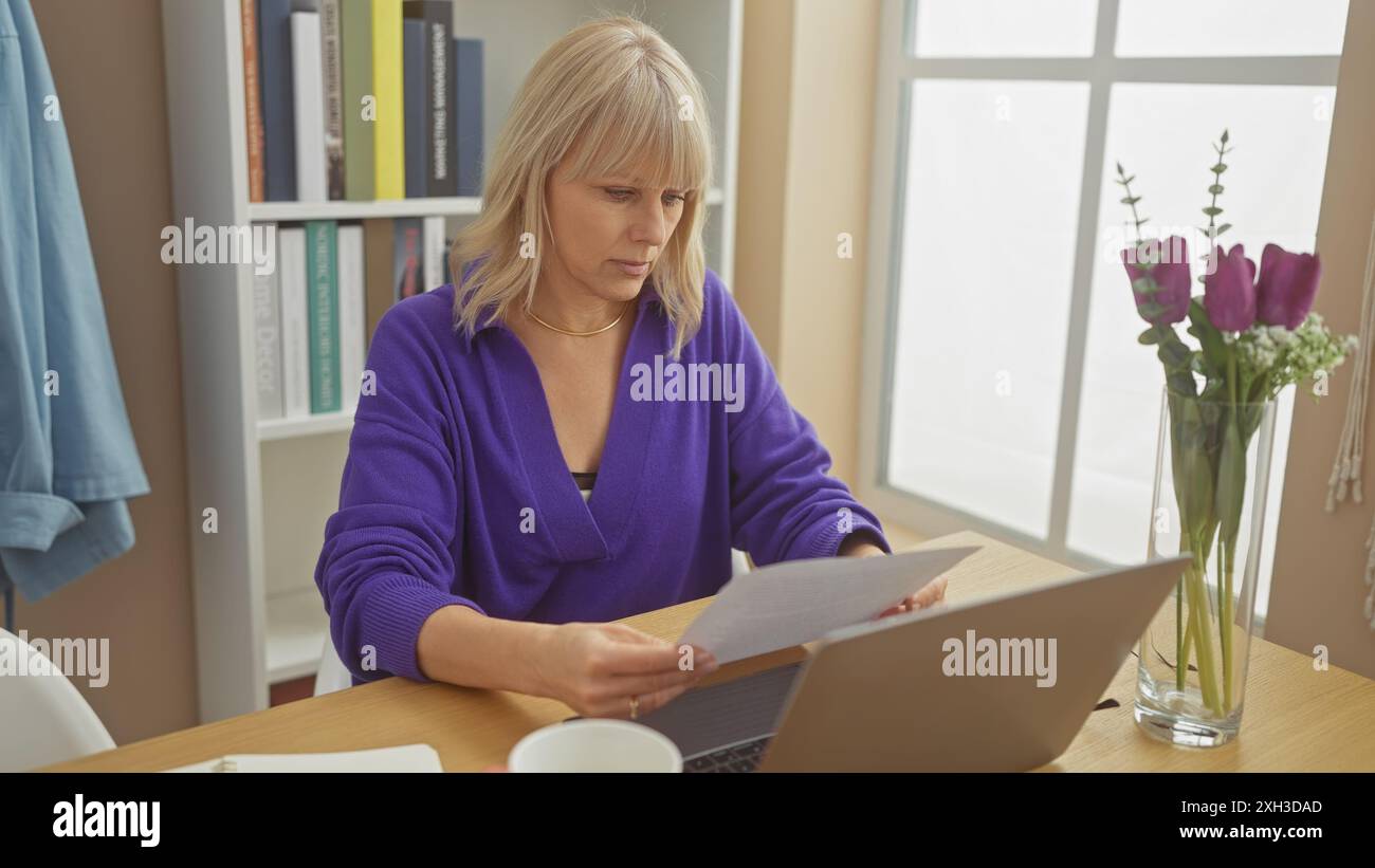 Blonde woman reading at home with laptop, flowers on table ...