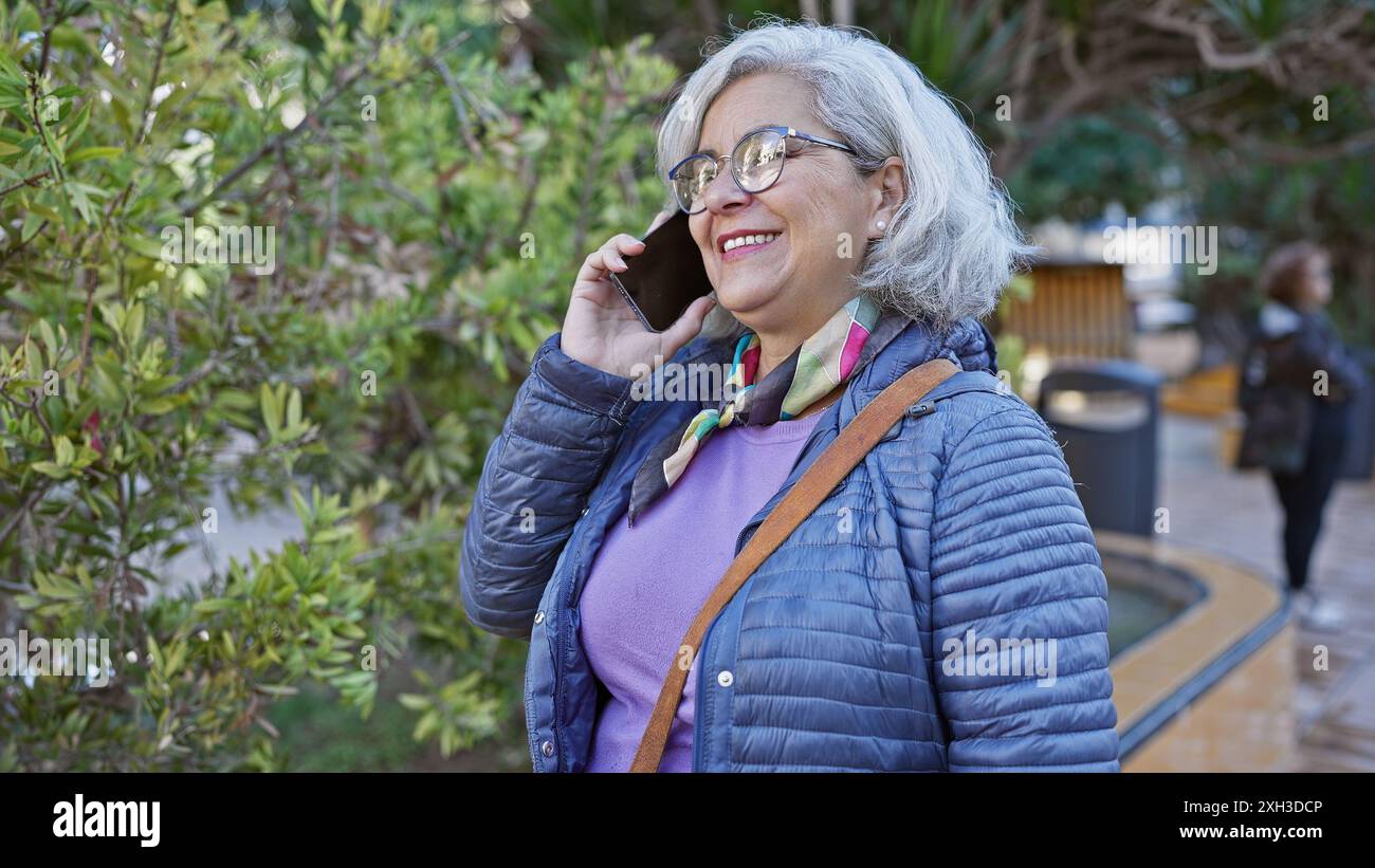 A smiling mature woman talks on a phone in a park surrounded by green ...