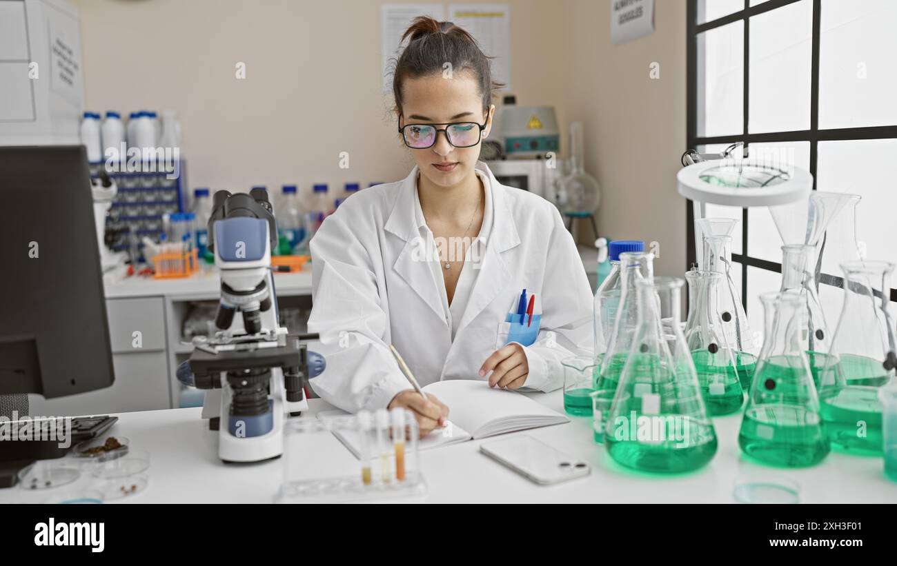 Focused hispanic woman scientist writing in notebook at laboratory workspace with microscope and ...