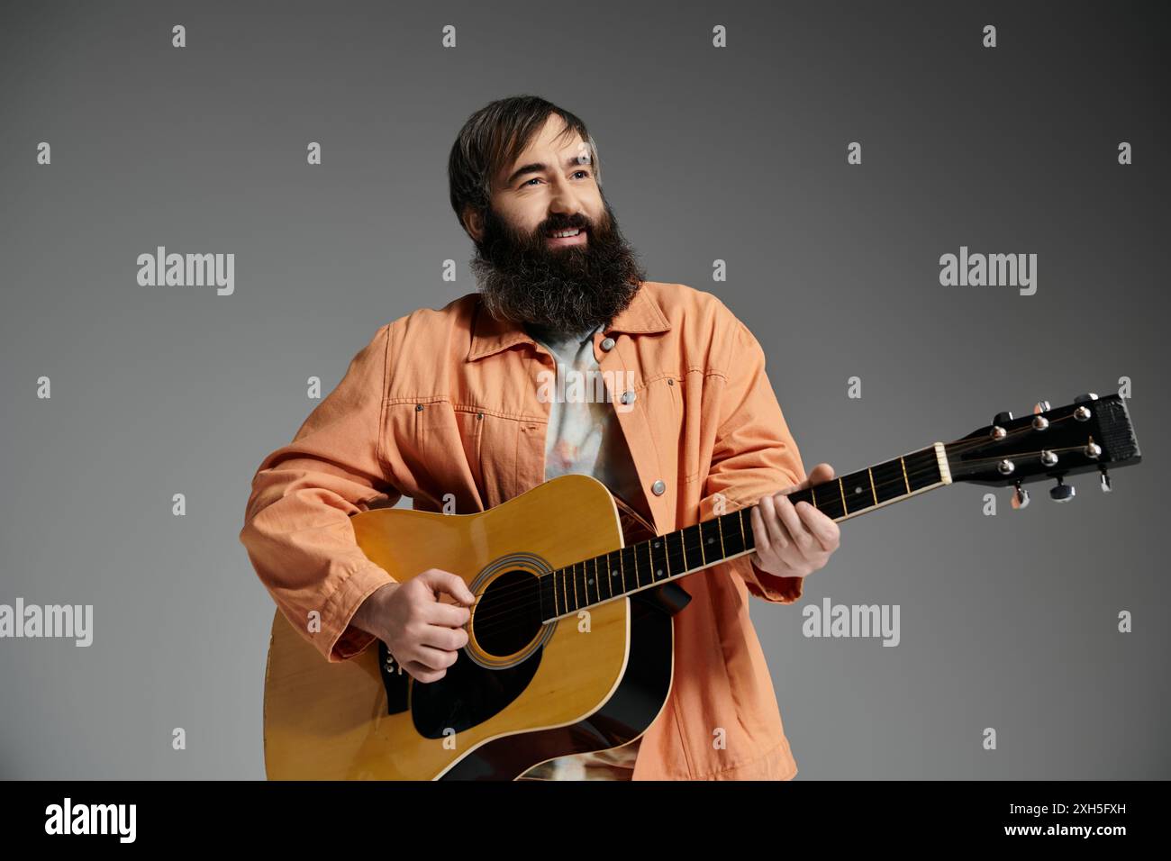A man with a beard plays an acoustic guitar in a studio with a grey ...