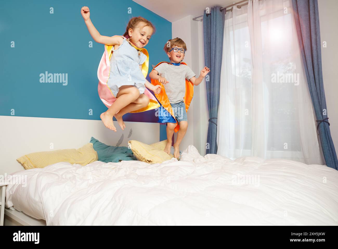 Two children wearing colorful capes are joyfully jumping on a bed in a ...
