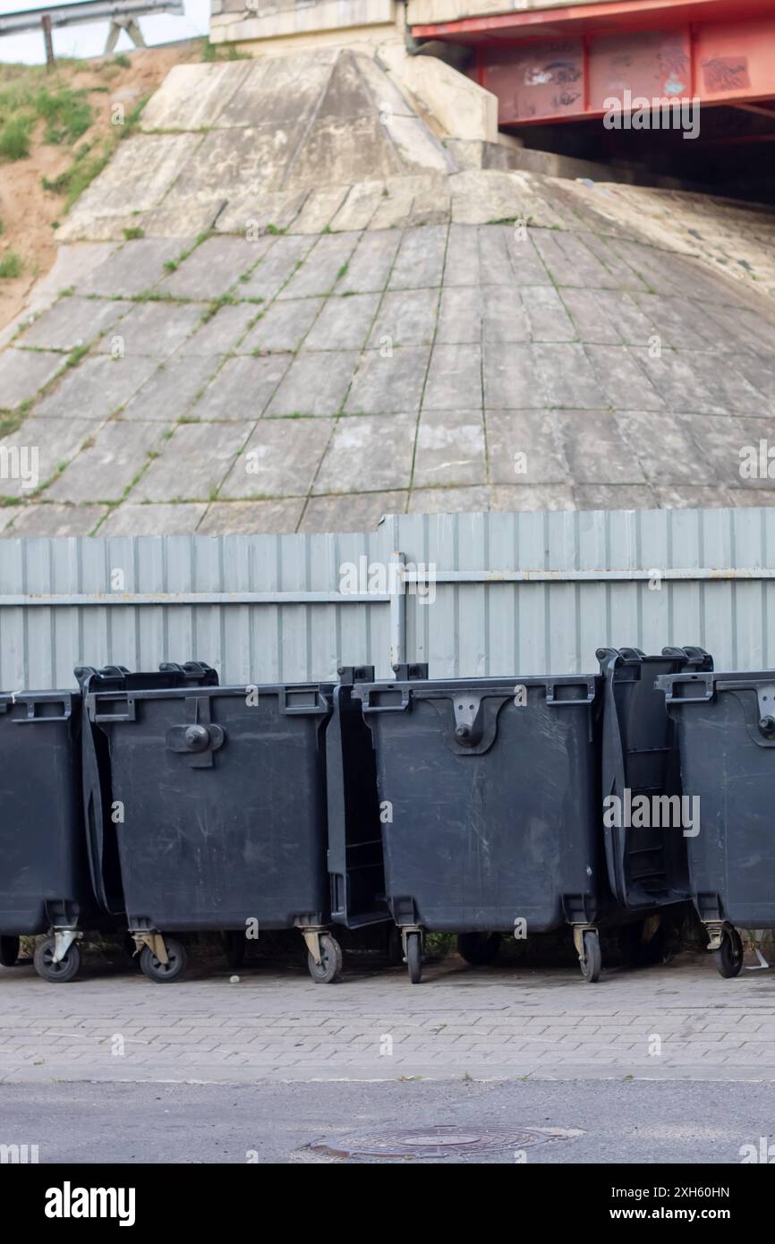 A row of black waste containers are placed in front of a wall, on ...