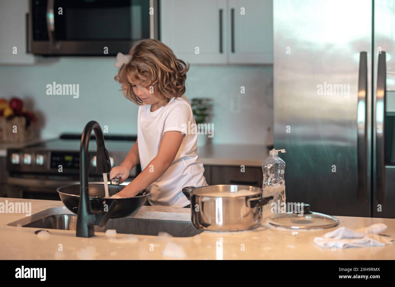 Kid washing dishes in the kitchen interior. Child helping his parents with housework. Kid boy ...
