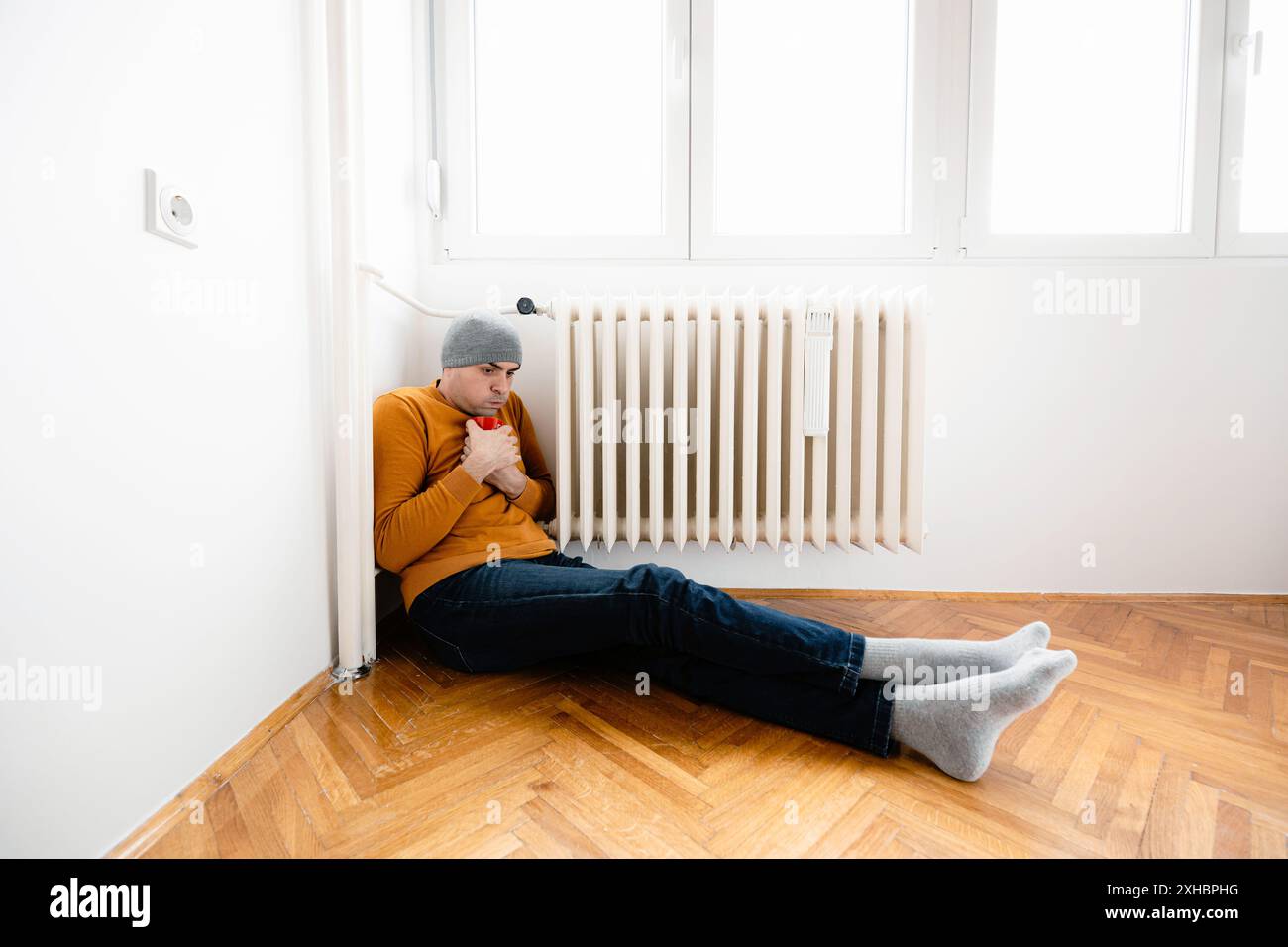 A man sits on the floor in a cold room, wearing a hat and an orange ...