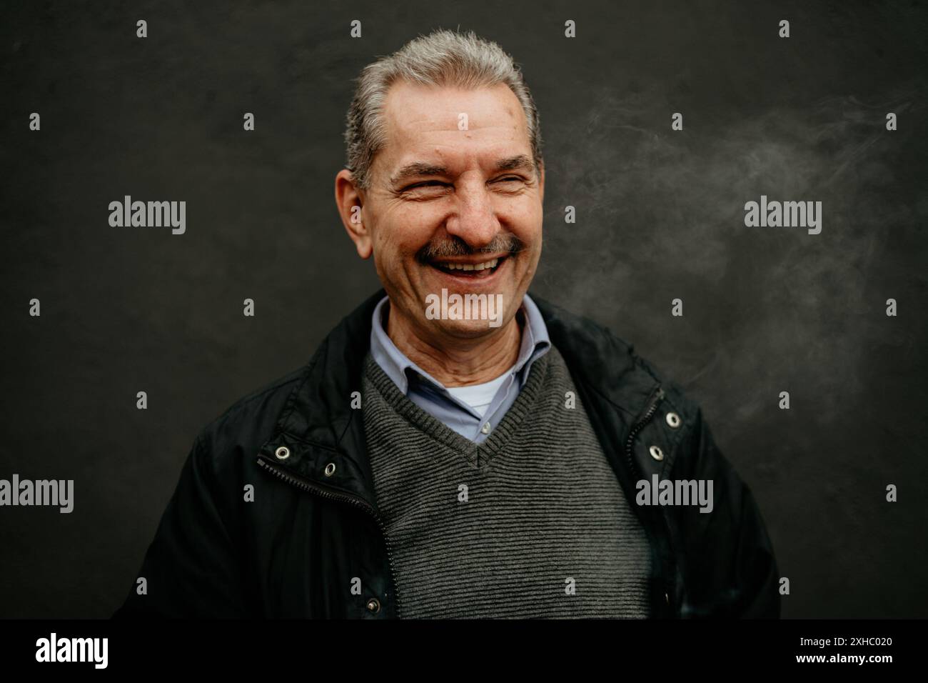 A man with grey hair and a mustache smiles broadly while wearing a black jacket over a grey ...