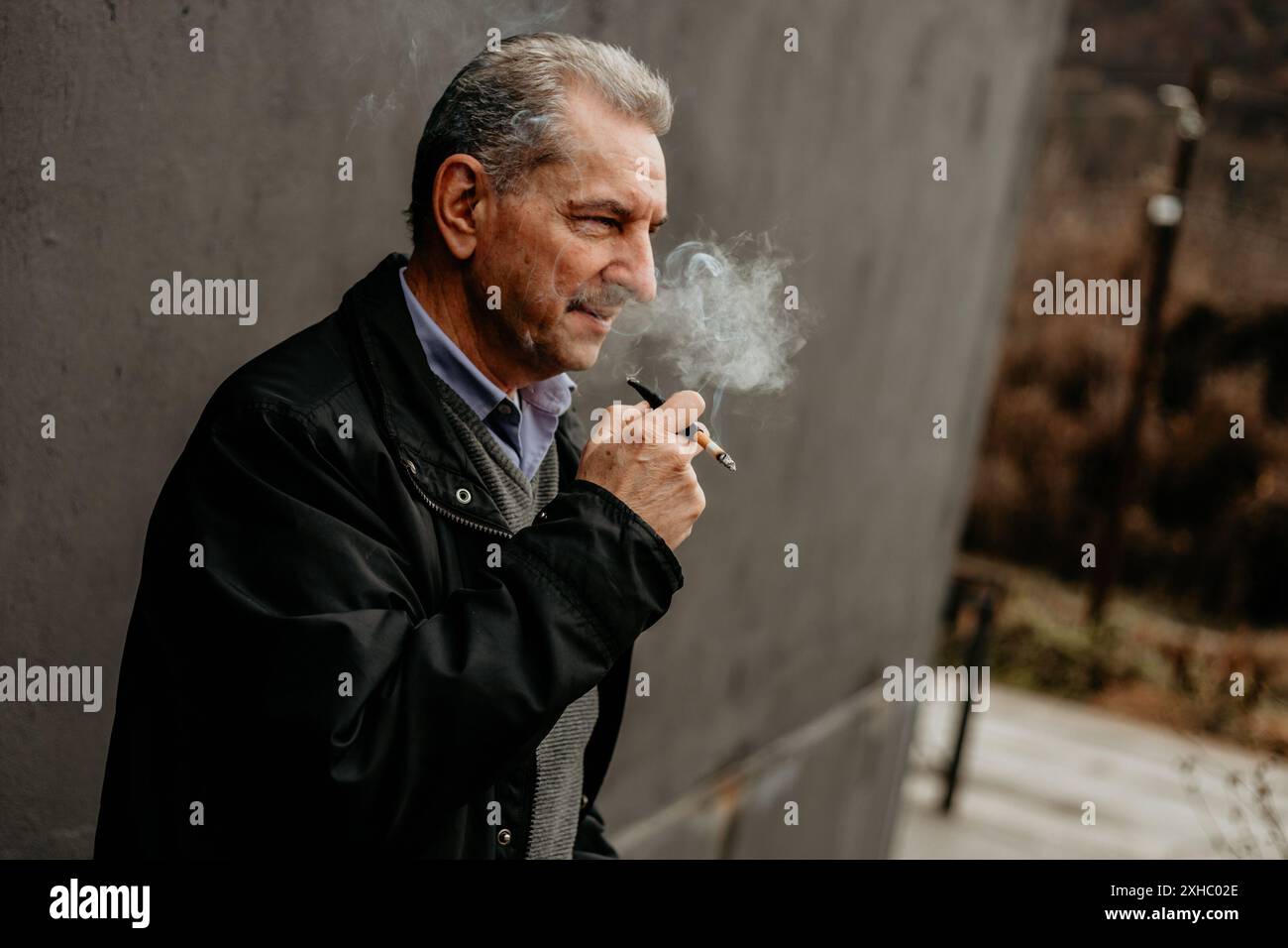 A man in a black jacket smokes a cigarette while standing against a ...