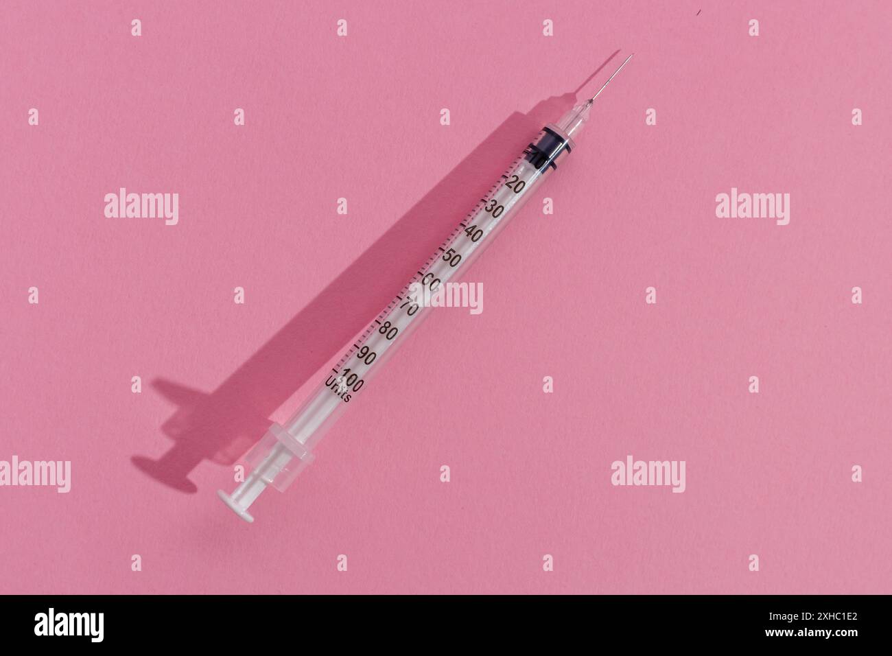 A clear syringe lies on a pink background, casting a shadow. The ...