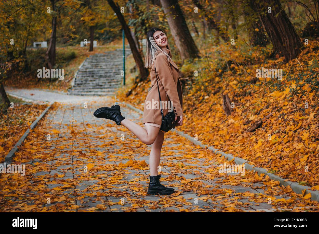 A woman walks down a path covered in fallen leaves, smiling and lifting ...