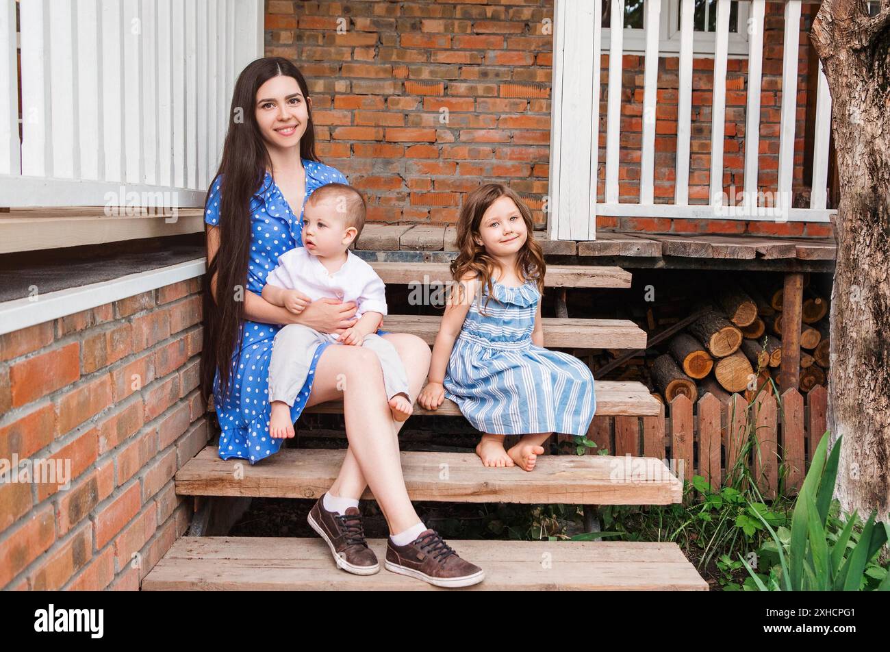 Young delighted mother sitting with adorable little siblings on terrace ...