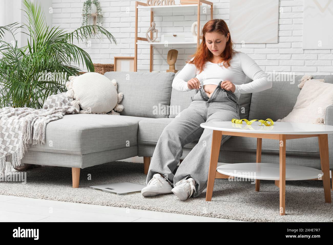 Sad overweight woman in tight jeans sitting on sofa at home. Excess ...