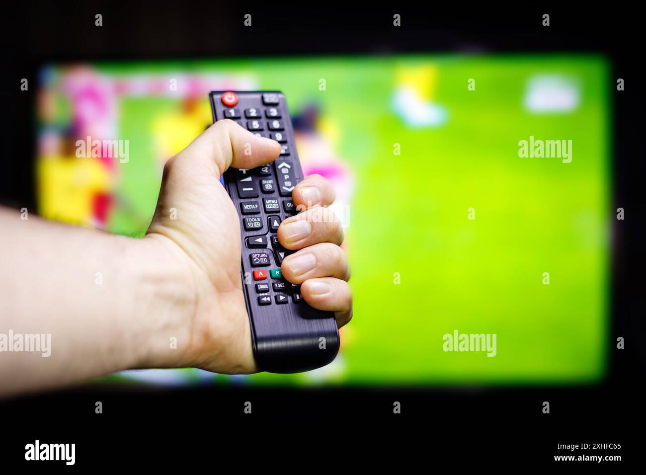 A hand holds a black TV remote control in front of a television screen ...