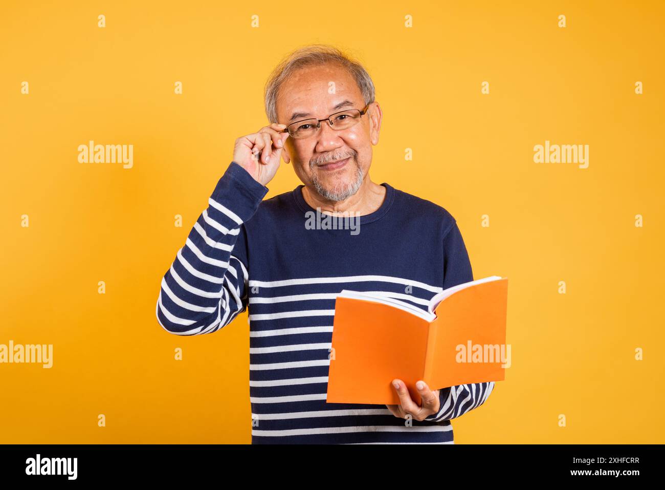 Portrait smiling Asian old man wearing glasses reading a book studio shot isolated yellow ...