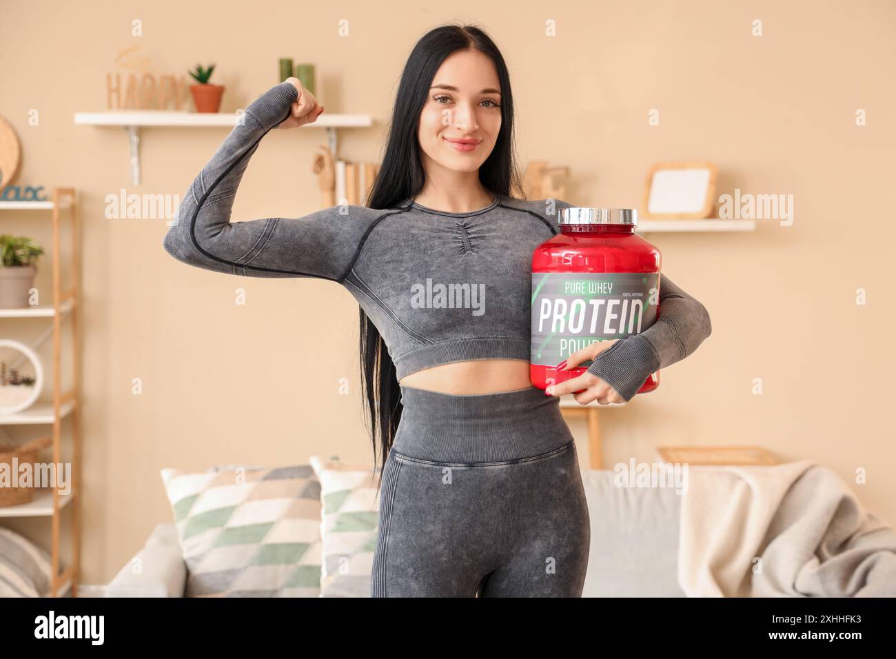 Sporty young woman with jar of protein powder showing muscles at home ...