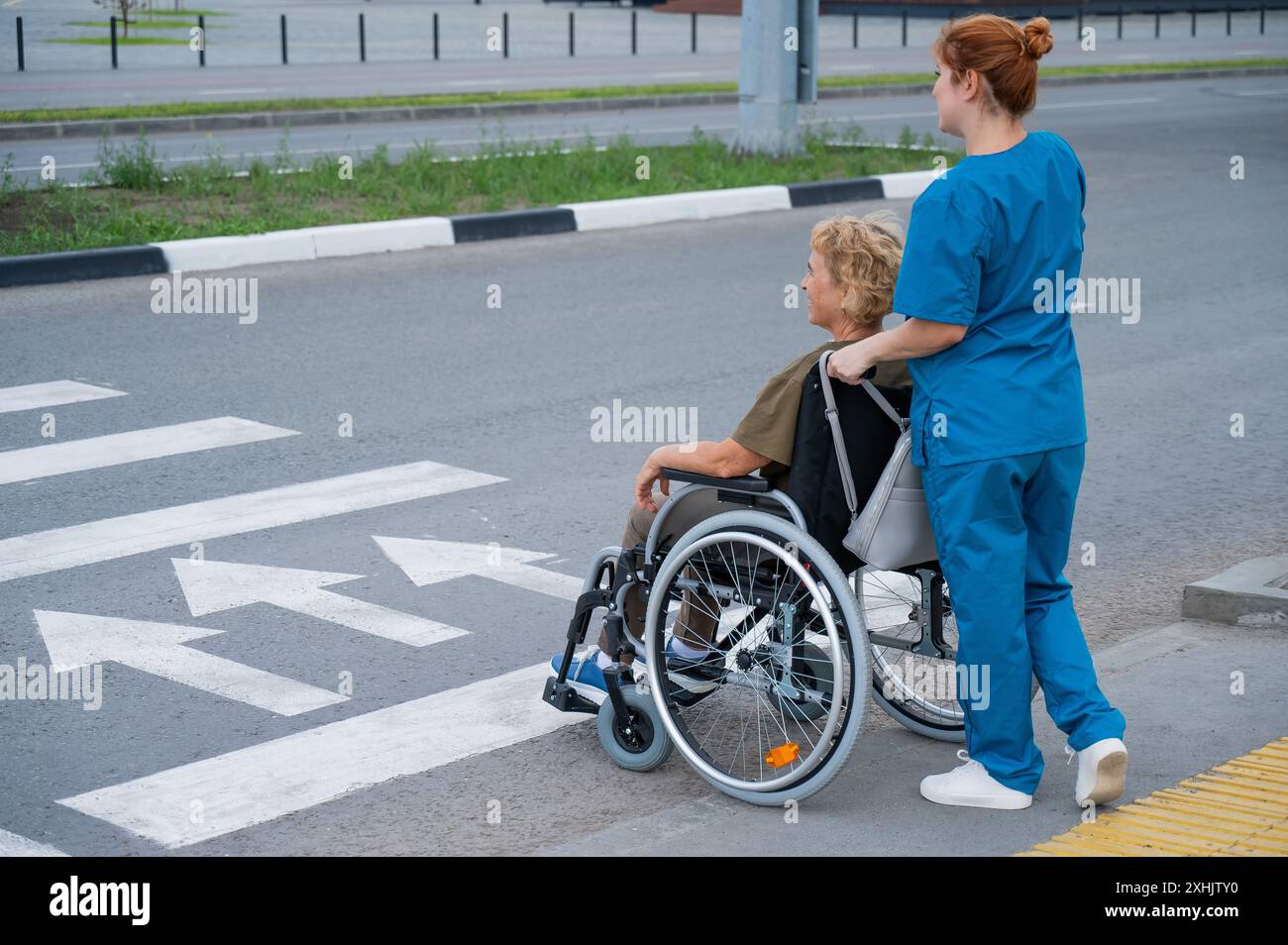 Rear view of a nurse helping an elderly woman in a wheelchair cross the ...
