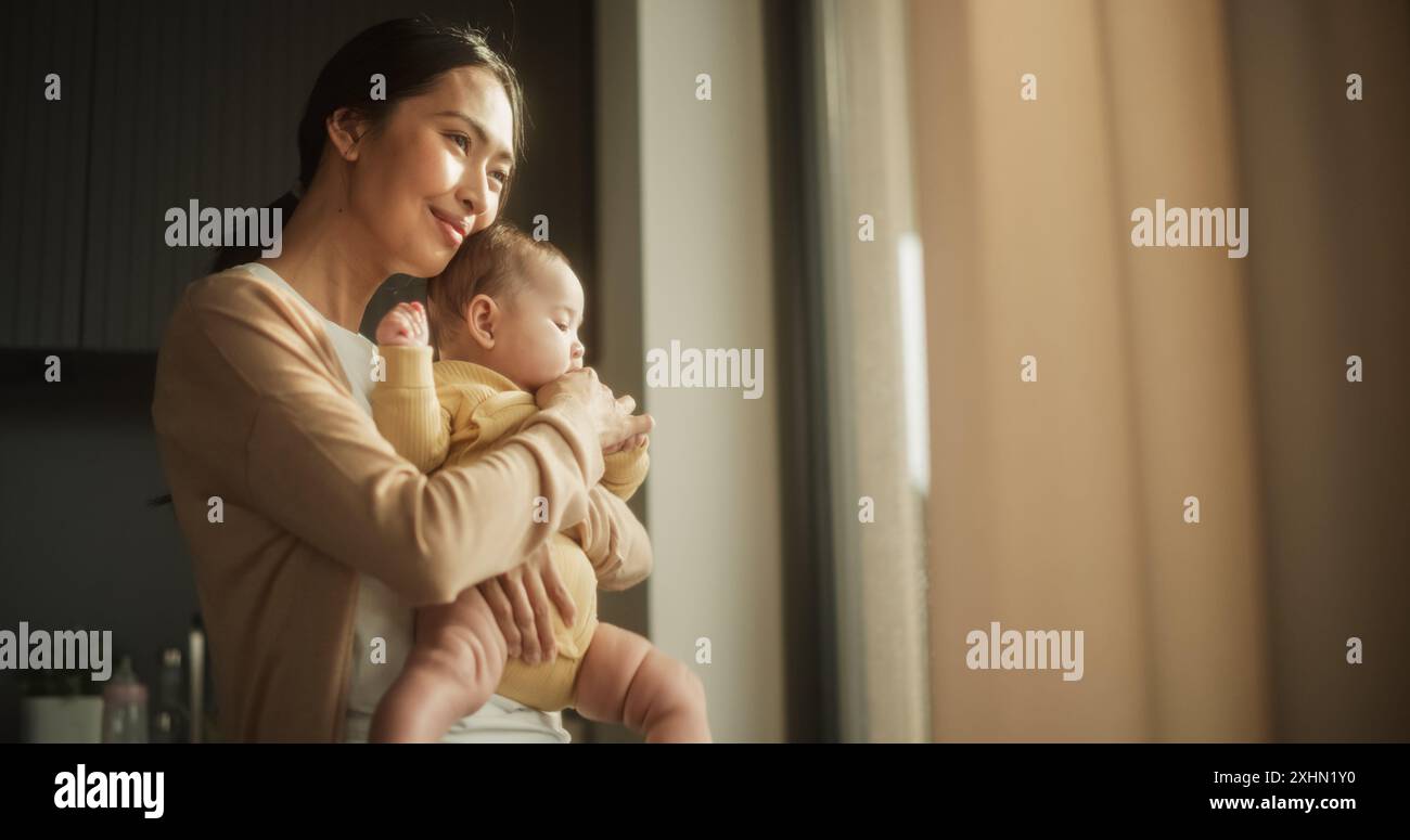 Beautiful Young Asian Mother Holding her Baby in her Arms While Standing Next to a Window at ...