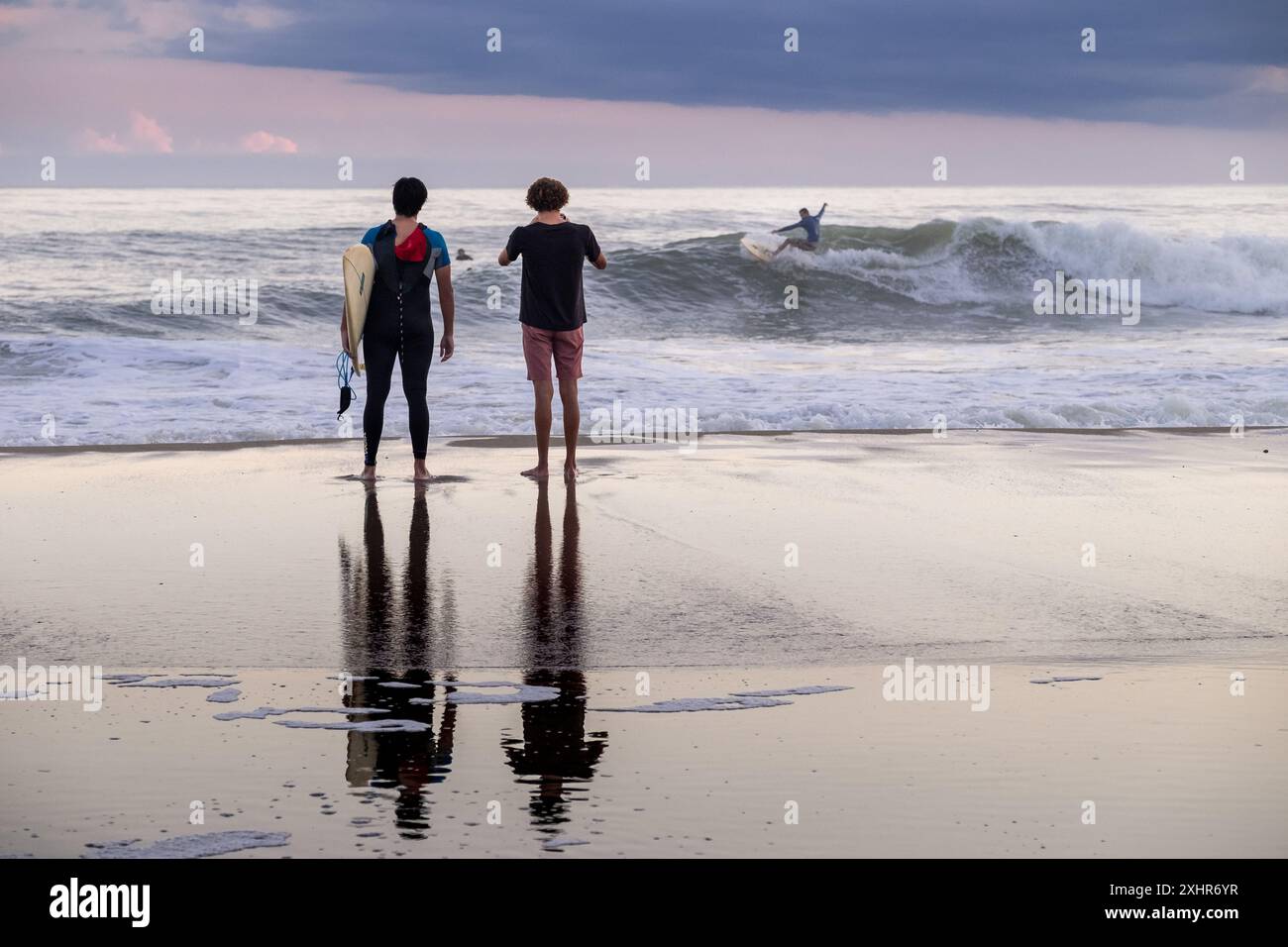 Two men standing on the beach, one holding a surfboard. The other man ...