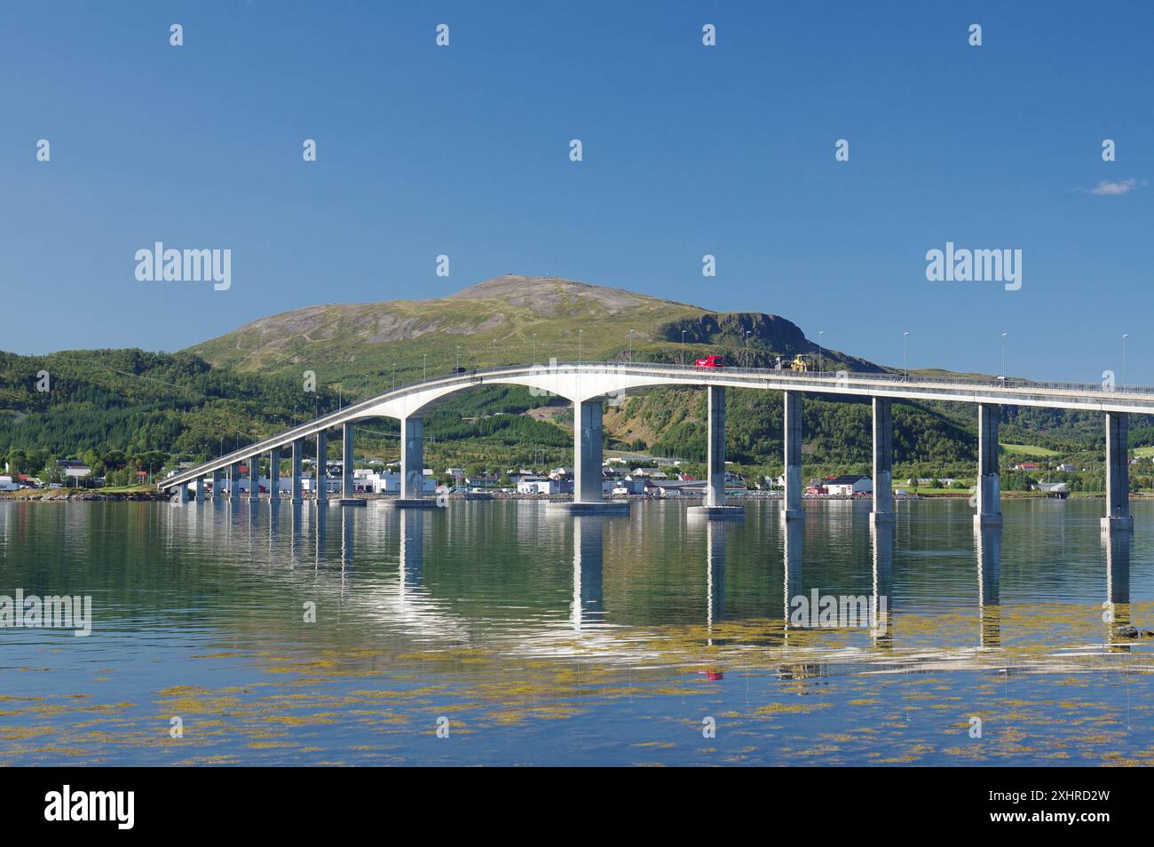 A curved bridge spans calm waters with reflective mountains and green ...