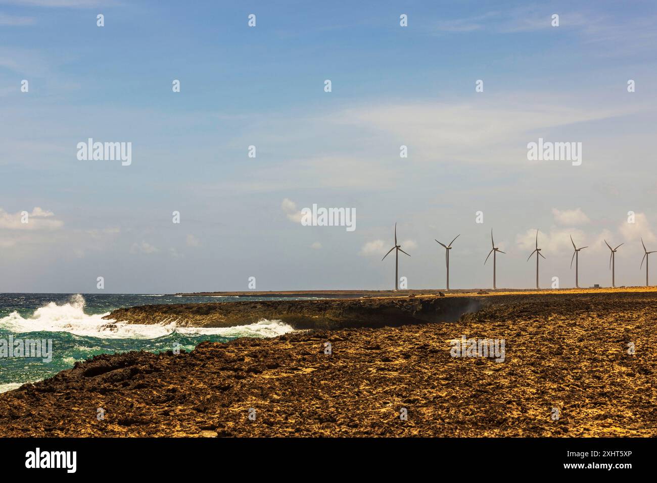 Wind turbines along rocky coastline with ocean waves and blue sky in ...