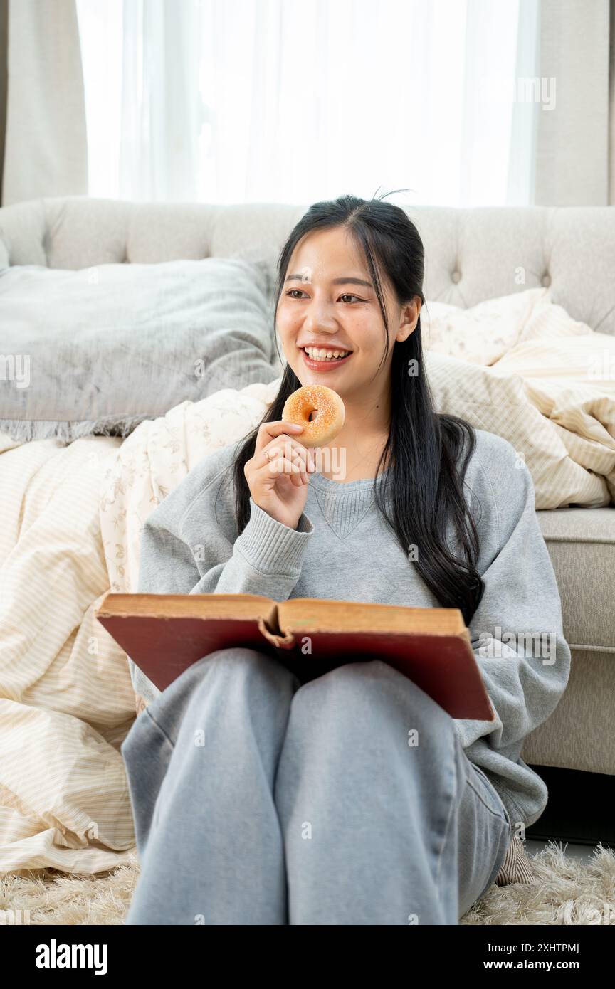 A happy, beautiful young Asian woman in comfy clothes is eating a donut ...