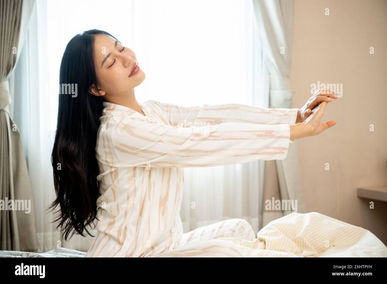 A beautiful, happy Asian woman in pajamas is sitting on the bed, keeping her eyes closed and ...