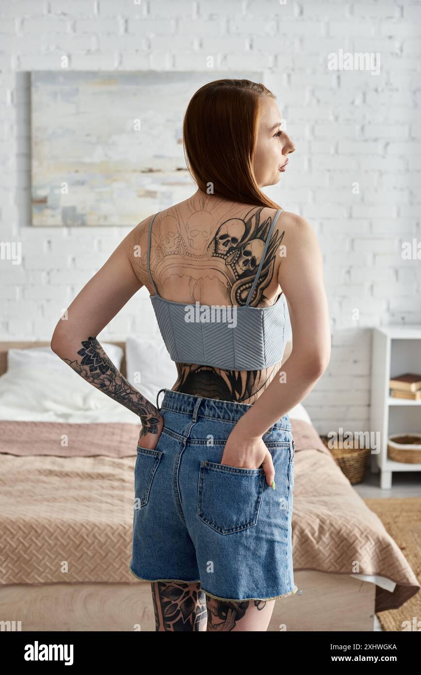 A young woman with tattoos on her back and arms stands in her modern ...